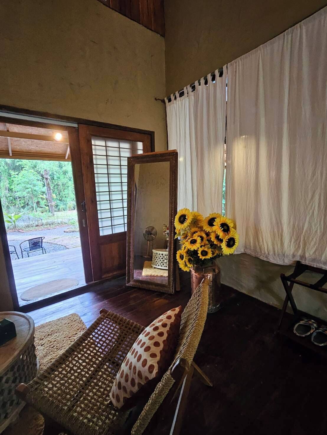 Um quarto com piso de madeira, uma cadeira de fibra natural, um espelho, um vaso com girassóis e vista para o exterior através de uma porta de correr.