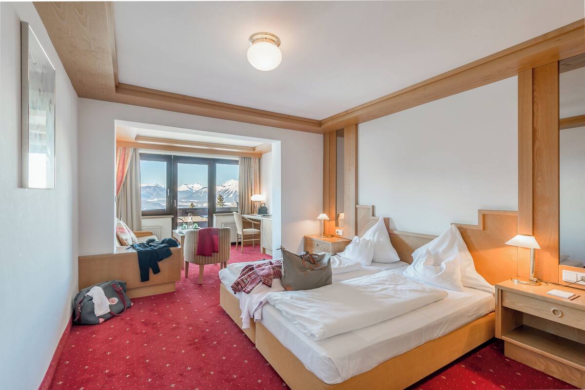 Quarto de hotel com cama, mesa de cabeceira, luminária e vista para montanhas pela janela.