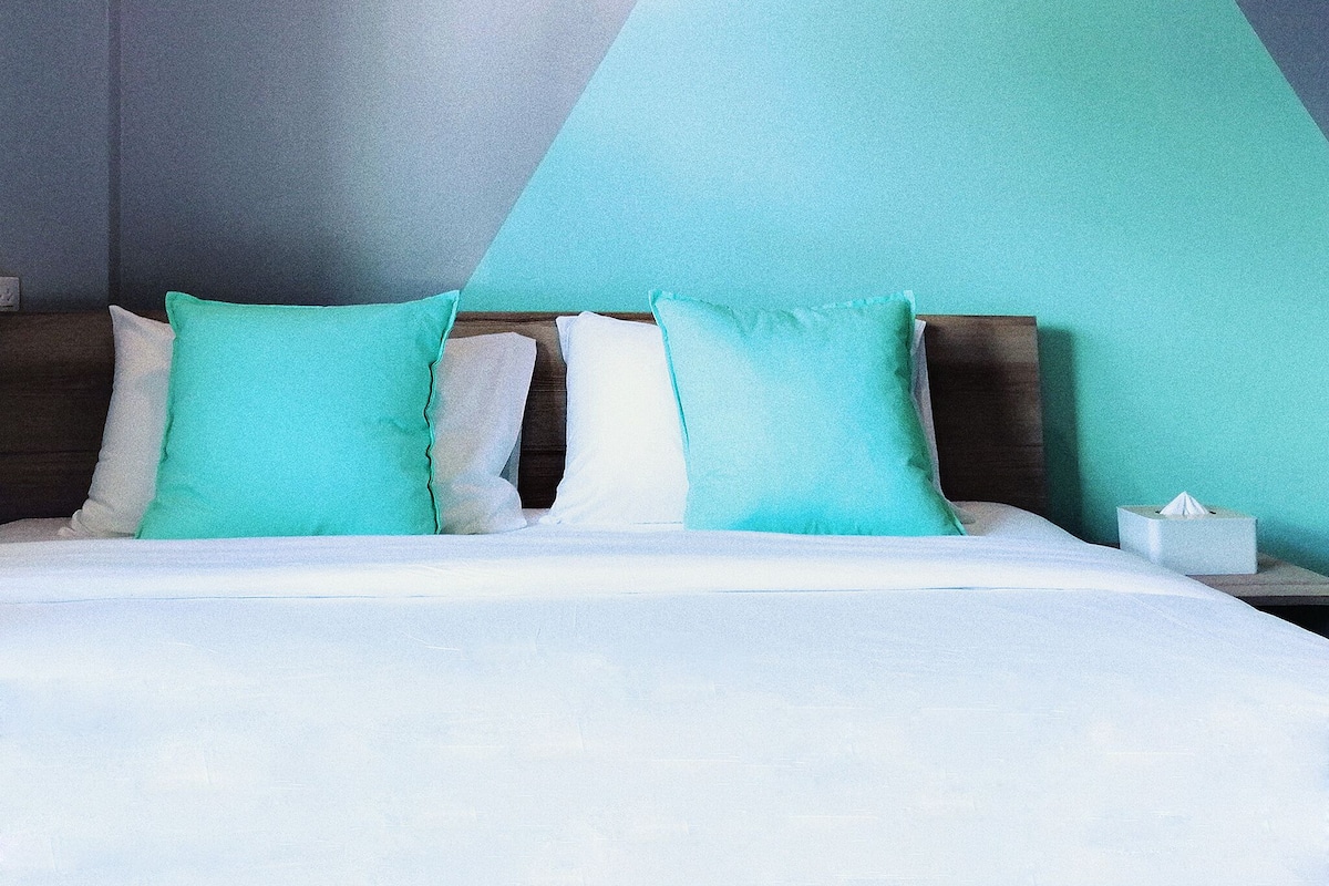 Cama com roupa de cama branca e almofadas verde-água, cabeceira de madeira e parede com padrão geométrico.