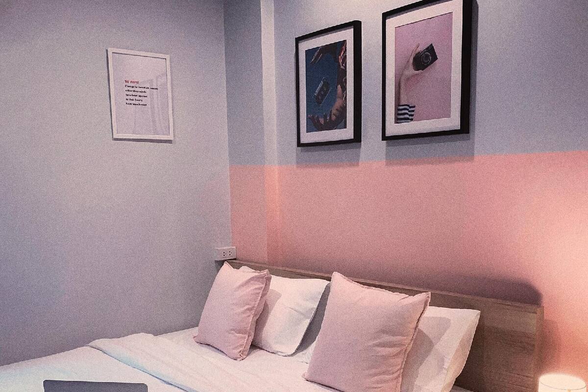 Cama bem arrumada com almofadas, um laptop sobre a cama e uma mesa de cabeceira com abajur. Há quadros emoldurados na parede.