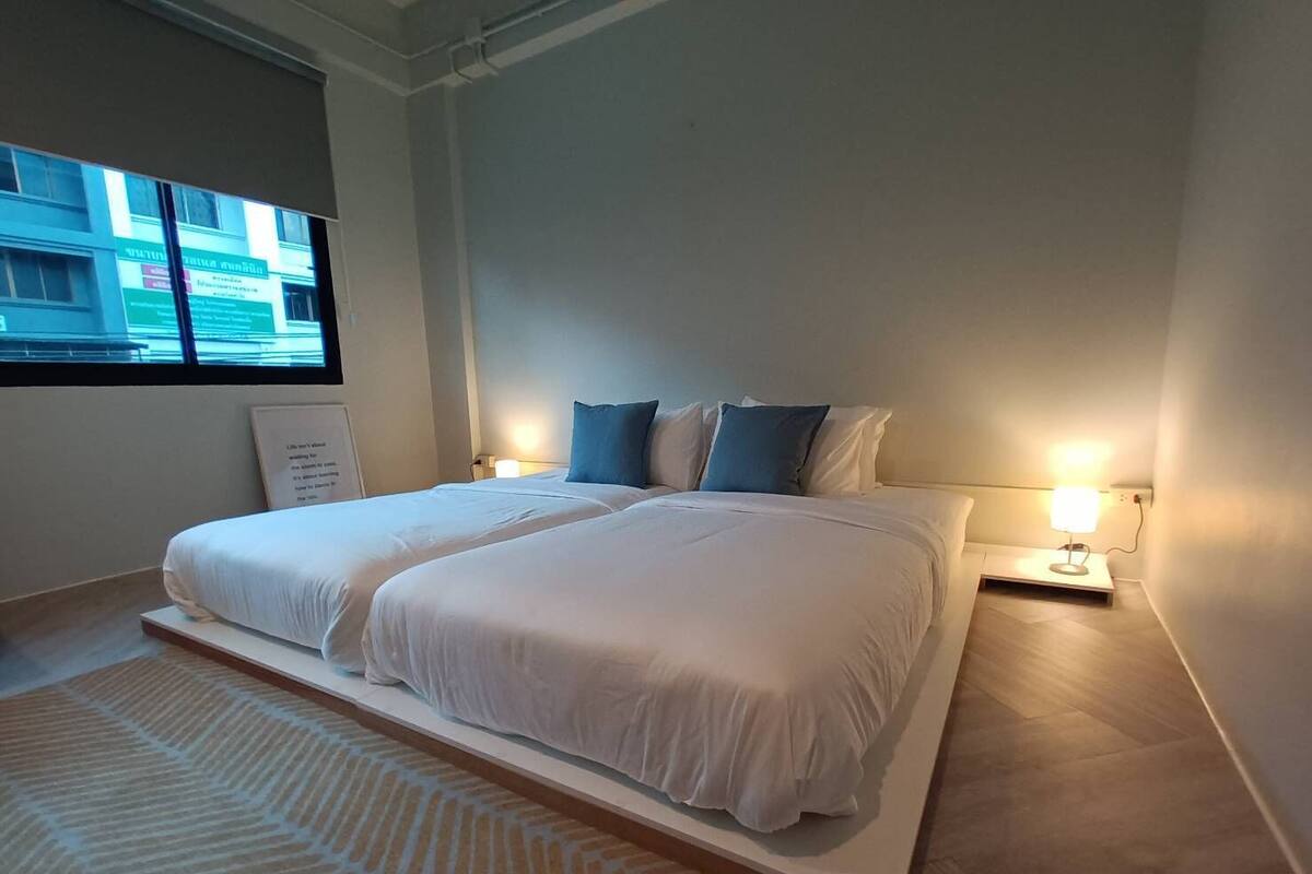 Uma cama de casal com roupa de cama branca e almofadas azuis, uma mesa de cabeceira com luminária e uma janela com vista para prédios.
