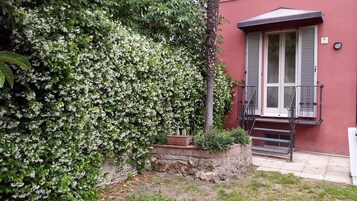 Um edifício vermelho com uma janela branca, um vaso de pedra com plantas e uma pequena escadaria que leva a uma varanda.