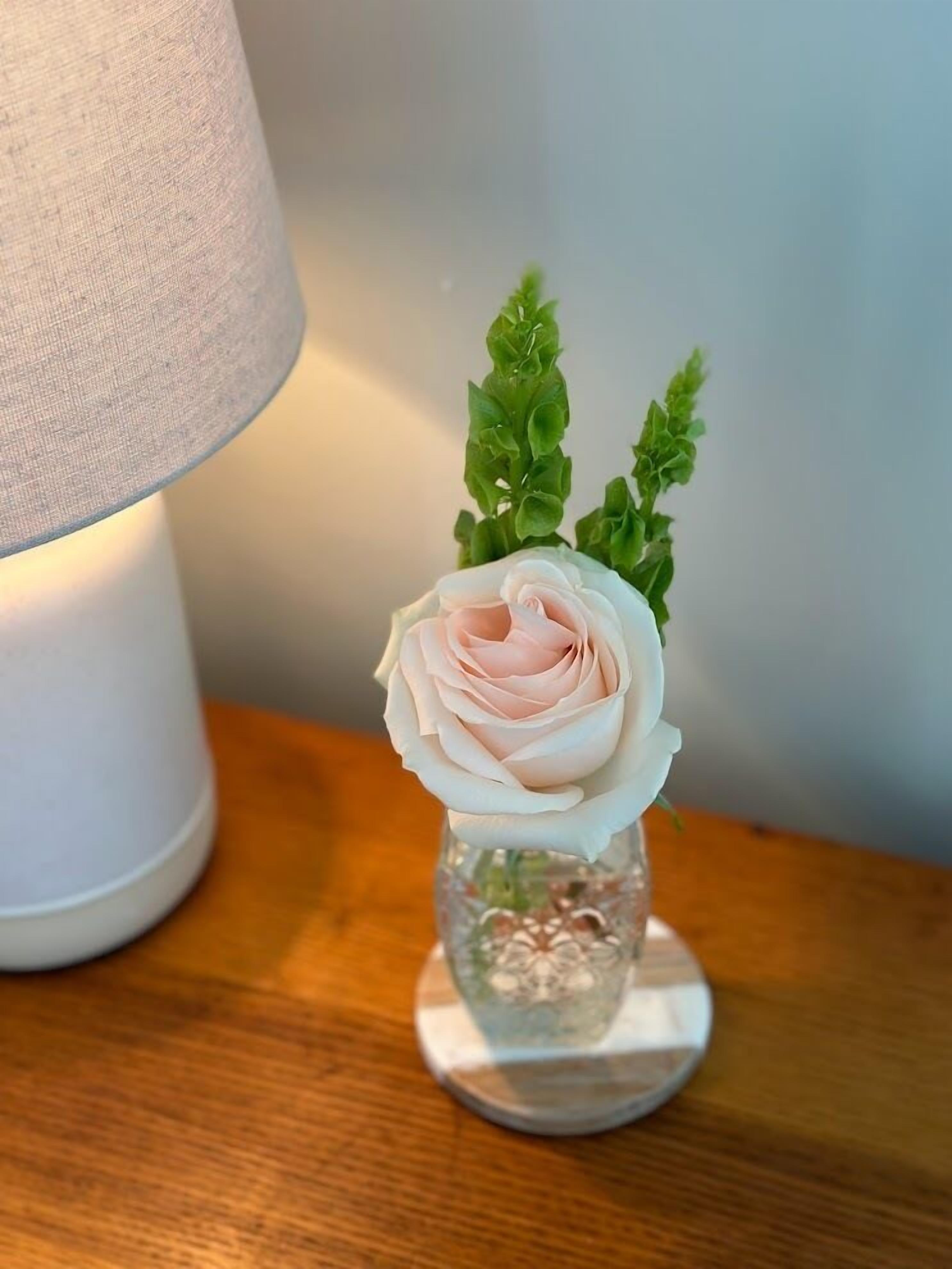 Um pequeno vaso de vidro com uma rosa rosa e folhagem verde sobre uma superfície de madeira.