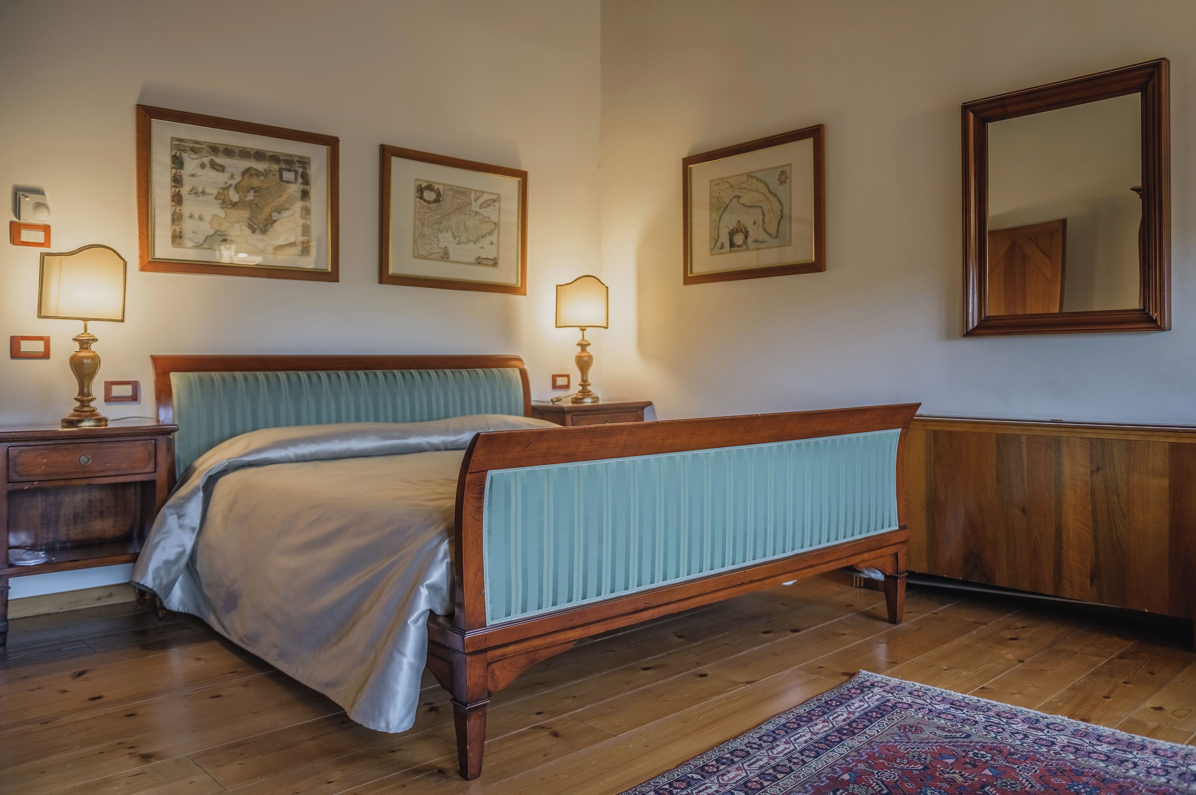 Quarto com cama de madeira, duas luminárias de cabeceira, mesa de cabeceira, cômoda e três mapas emoldurados na parede.