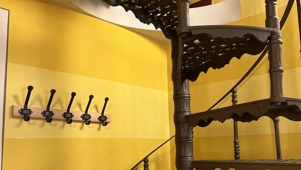 Escadaria em espiral com grades de metal ricamente decoradas, em frente a uma parede amarela.