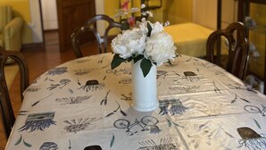 Uma mesa de jantar com toalha florida, um vaso branco com flores e uma escada ao fundo.