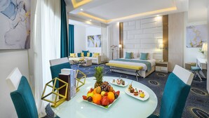 Quarto de hotel moderno com uma cama, um espaço de estar com uma mesa de centro e uma mesa de jantar com frutas e doces.