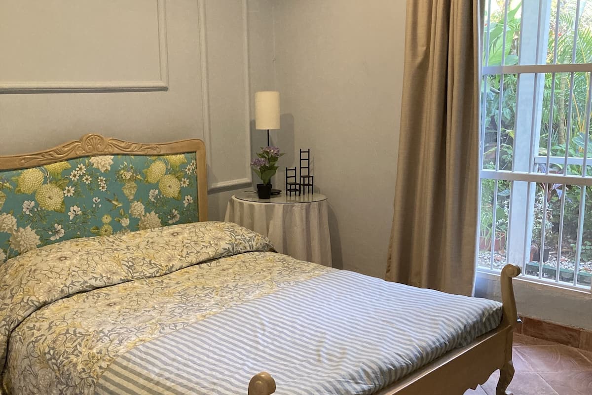 Um quarto com colcha de padrão floral, estrutura de cama de madeira, uma mesinha redonda com abajur e uma janela com cortinas.