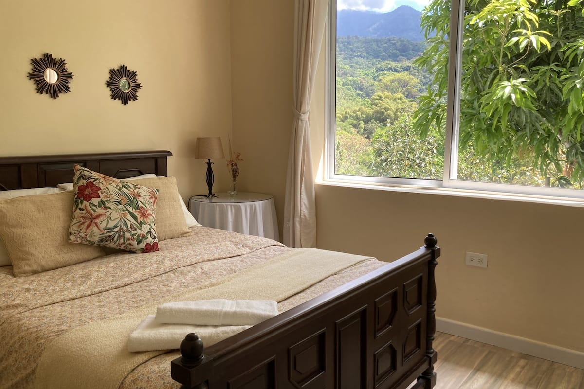 Quarto com cama, mesa de cabeceira, luminária e janela com vista para o verde.