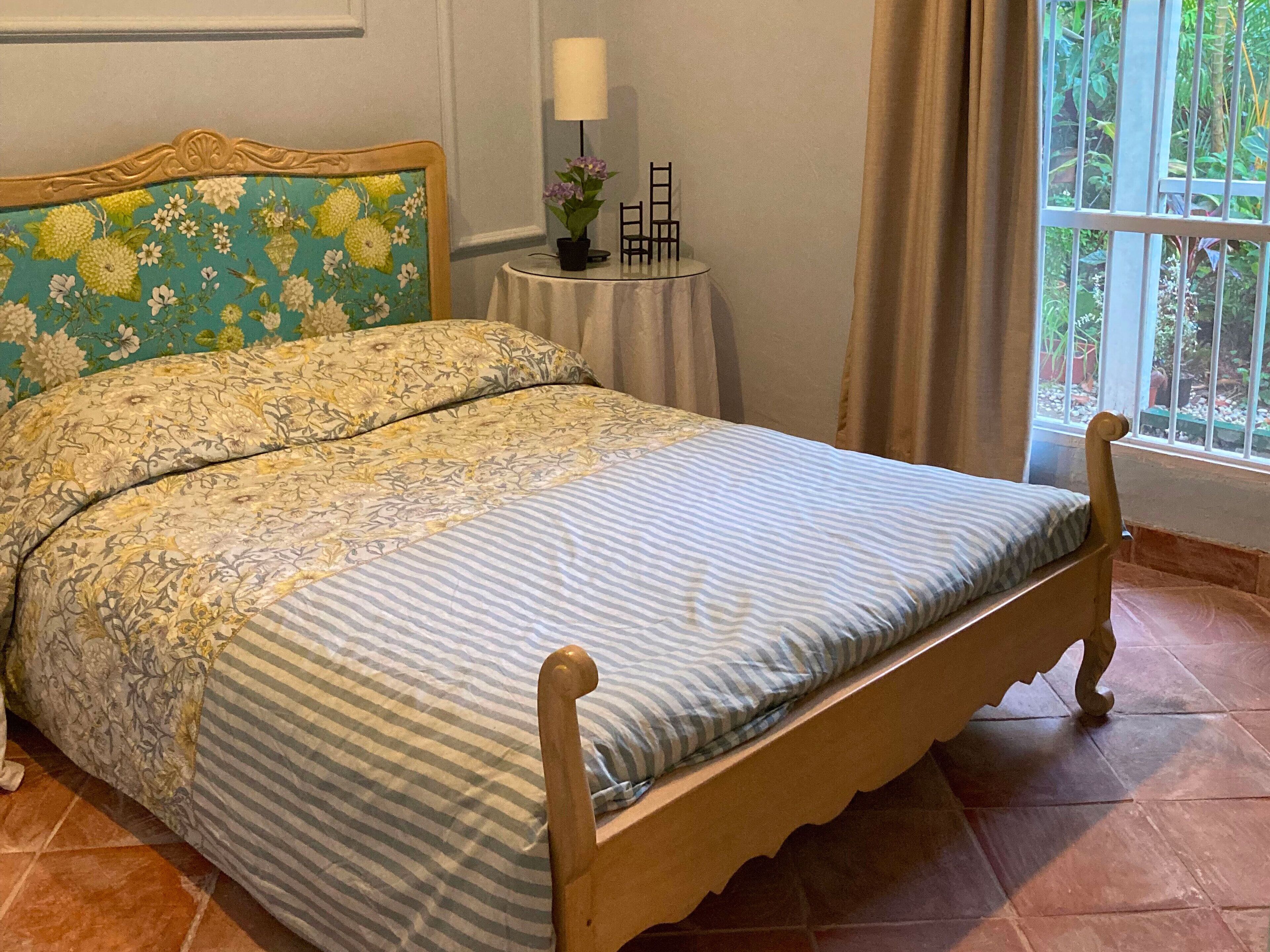 Cama com cabeceira floral e roupa de cama listrada, uma mesinha com abajur e uma janela com cortinas.
