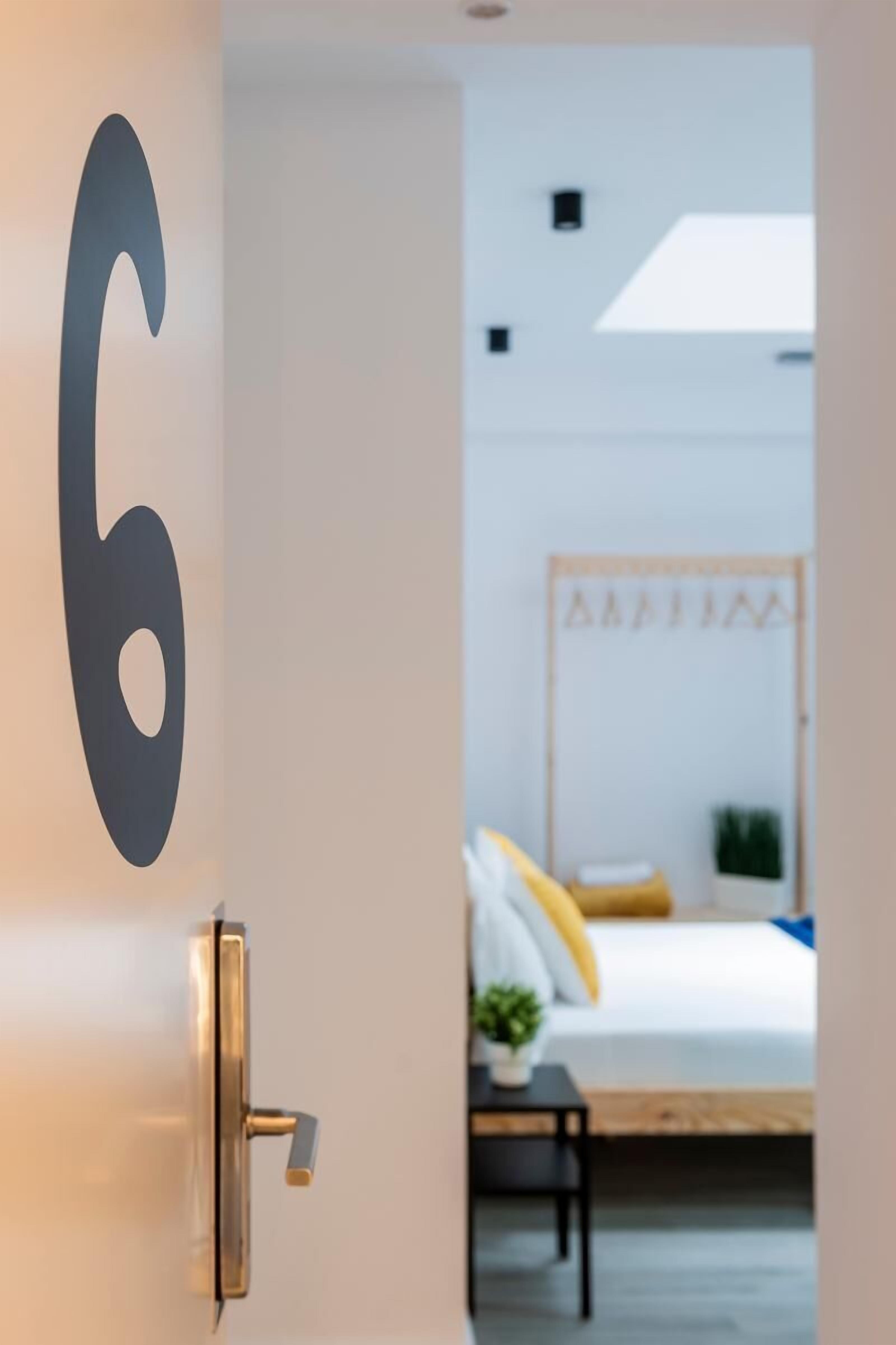 Uma porta com o número 6, que dá para um quarto com uma cama, uma mesinha de cabeceira e uma pequena planta.