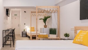Quarto de hotel com cama, escrivaninha, cadeira, cabideiro e planta em vaso.