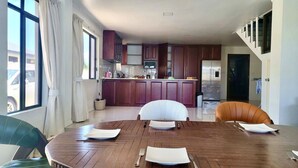 Uma área de refeições com mesa de madeira posta para quatro pessoas, cozinha ao fundo com armários de madeira escura e eletrodomésticos de aço inoxidável, e vista para carros estacionados lá fora.