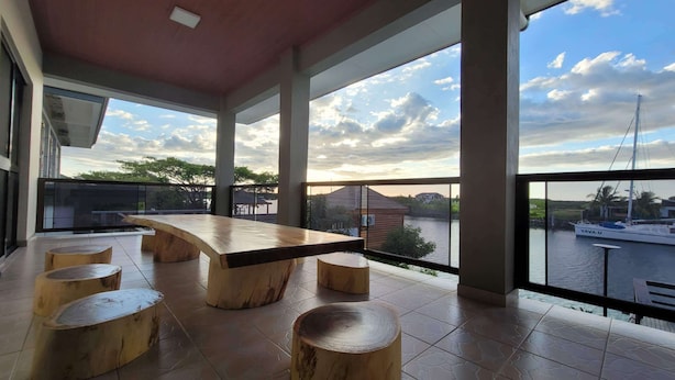 Uma varanda ampla com uma longa mesa de madeira e vários banquinhos redondos de madeira, com vista para um corpo d'água onde um barco está atracado.