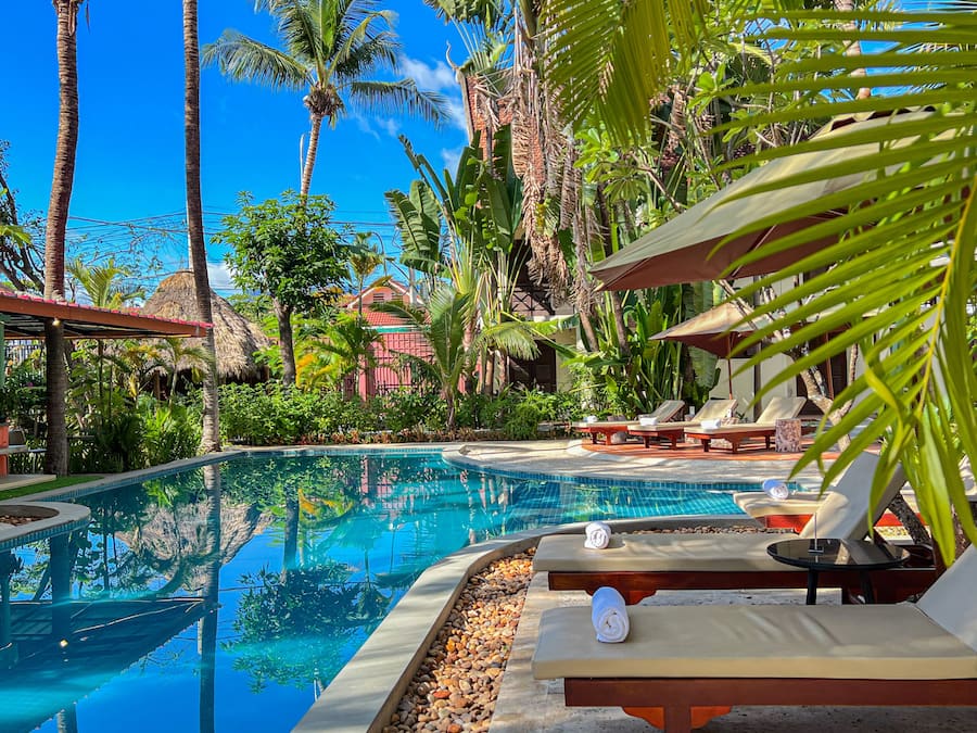 Uma área de piscina com espreguiçadeiras e guarda-sóis, cercada por palmeiras e plantas tropicais.