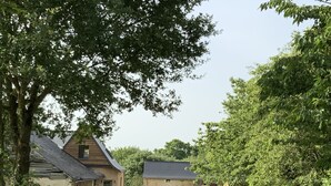 Um ambiente rural com dois edifícios de pedra, um com telhado inclinado e outro com telhado plano, rodeados por vegetação e um céu limpo.