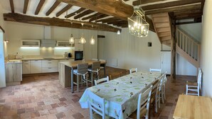 Uma cozinha rústica com vigas aparentes, um espaço de refeições com uma mesa comprida e cadeiras, e uma escadaria que leva a um andar superior.