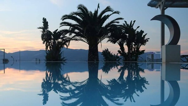 Área de piscina tranquila com palmeiras e um reflexo límpido na água, rodeada por montanhas.