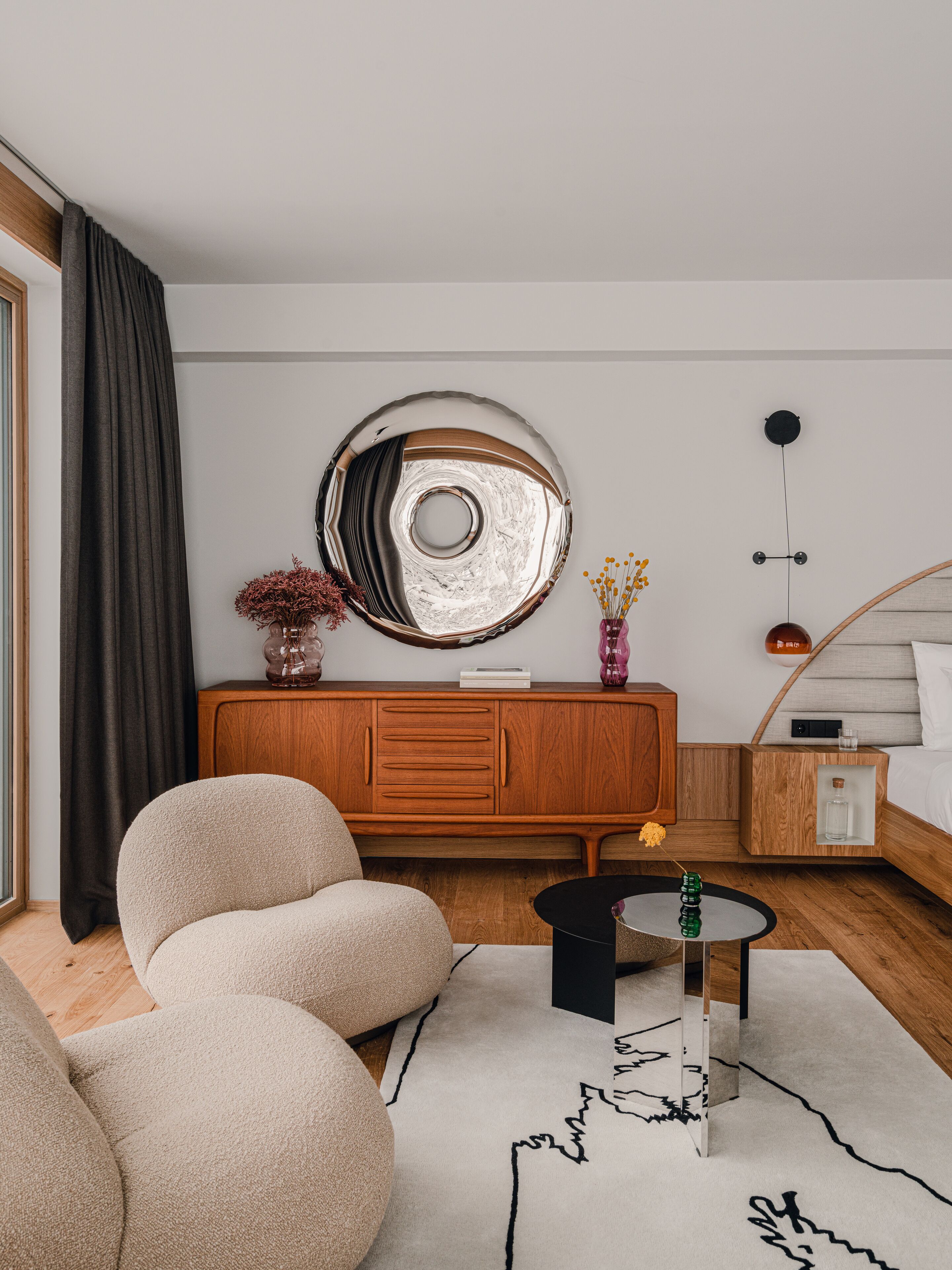 Quarto moderno com cômoda de madeira, espelho redondo, cama com cabeceira de madeira, mesa de centro redonda e sofá bege.