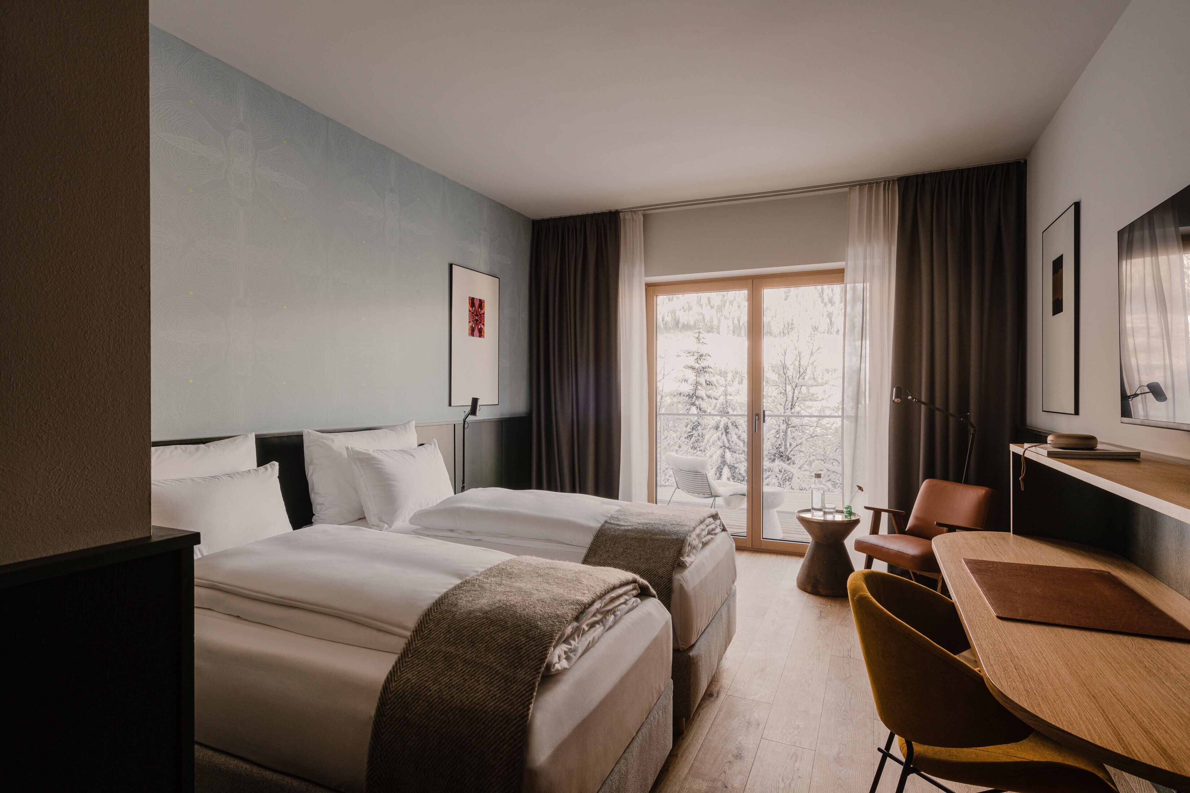Quarto de hotel com duas camas, uma escrivaninha e uma cadeira. Há uma janela com cortinas e vista para árvores.