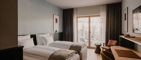 Quarto de hotel com duas camas, uma escrivaninha e uma cadeira. Há uma janela com cortinas e vista para árvores.