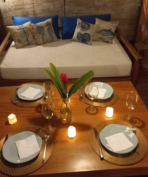 Uma mesa de jantar posta com pratos, talheres e velas, com um sofá e almofadas ao fundo.