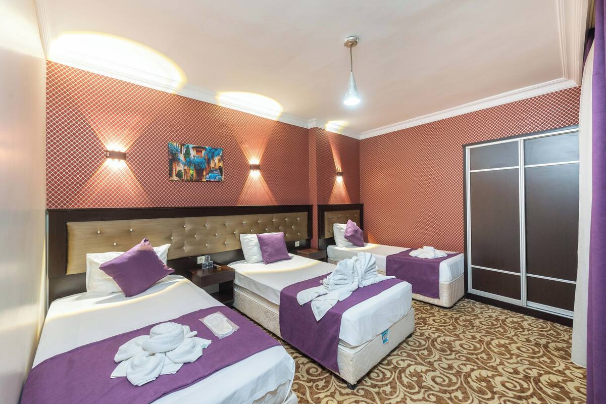 Quarto de hotel com duas camas, cada uma com um cobertor e toalhas roxas, uma grande obra de arte na parede e um armário com porta deslizante.