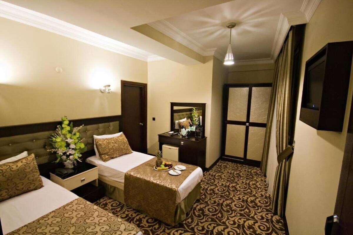 Quarto de hotel com duas camas, uma escrivaninha, um espelho, uma televisão e um tapete estampado.
