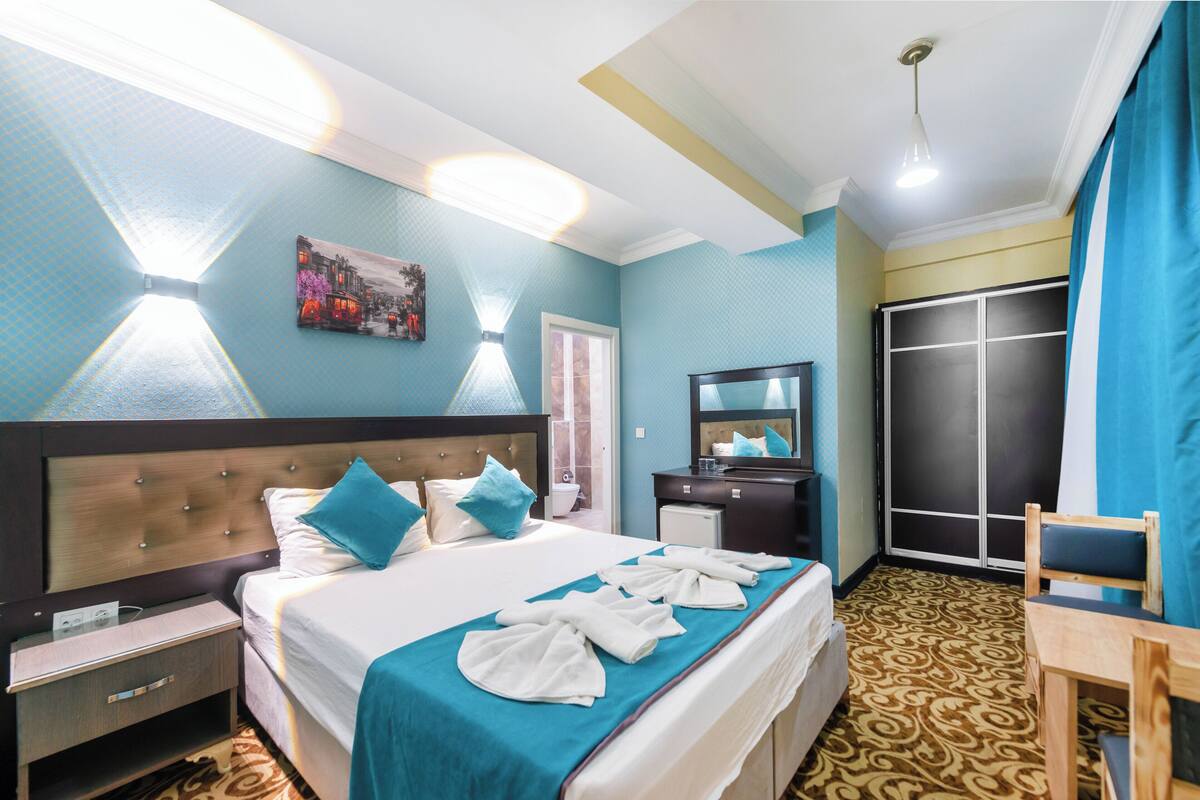 Quarto de hotel com cama, mesa de cabeceira, guarda-roupa e pequena escrivaninha com cadeira.