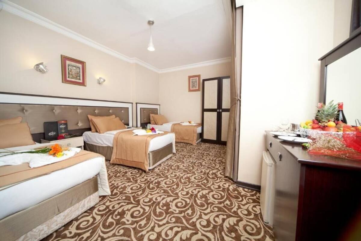 Quarto de hotel com duas camas, um minibar, uma mesa com fruta e uma garrafa, e um carpete estampado.