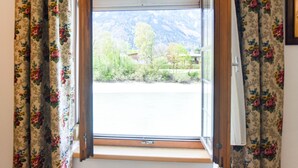 Uma janela com cortinas florais, vista de uma paisagem montanhosa e um edifício ao longe.