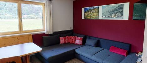 Um quarto aconchegante com uma mesa de madeira, um sofá cinza com almofadas vermelhas e uma janela com vista para o verde.