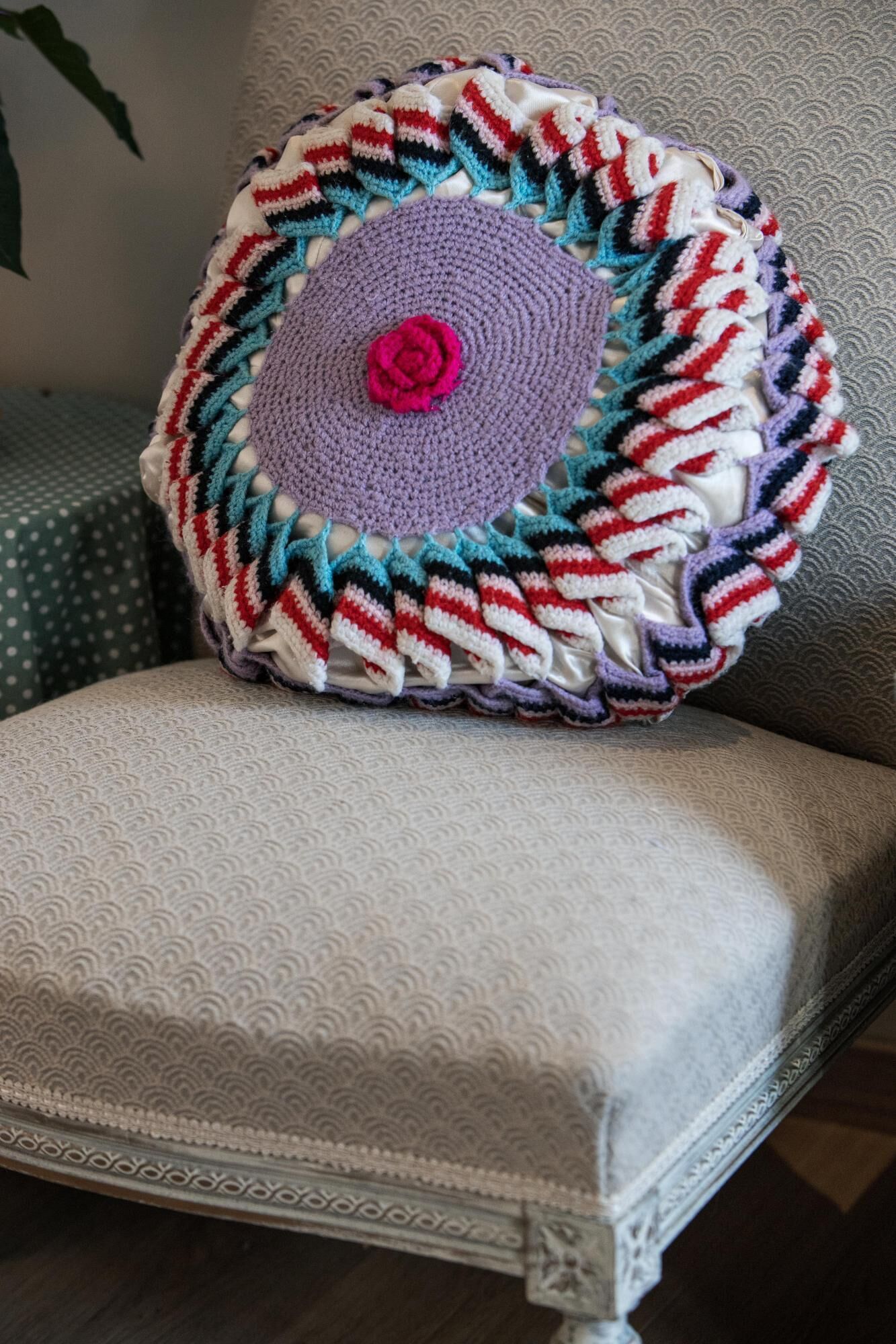 Um travesseiro de crochê com um desenho floral central, colocado sobre uma cadeira com almofada texturizada.