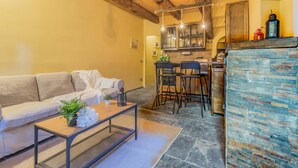 Uma sala de estar aconchegante com um sofá, uma mesa de centro com plantas e uma área de cozinha com bancada de pedra e banquetas.