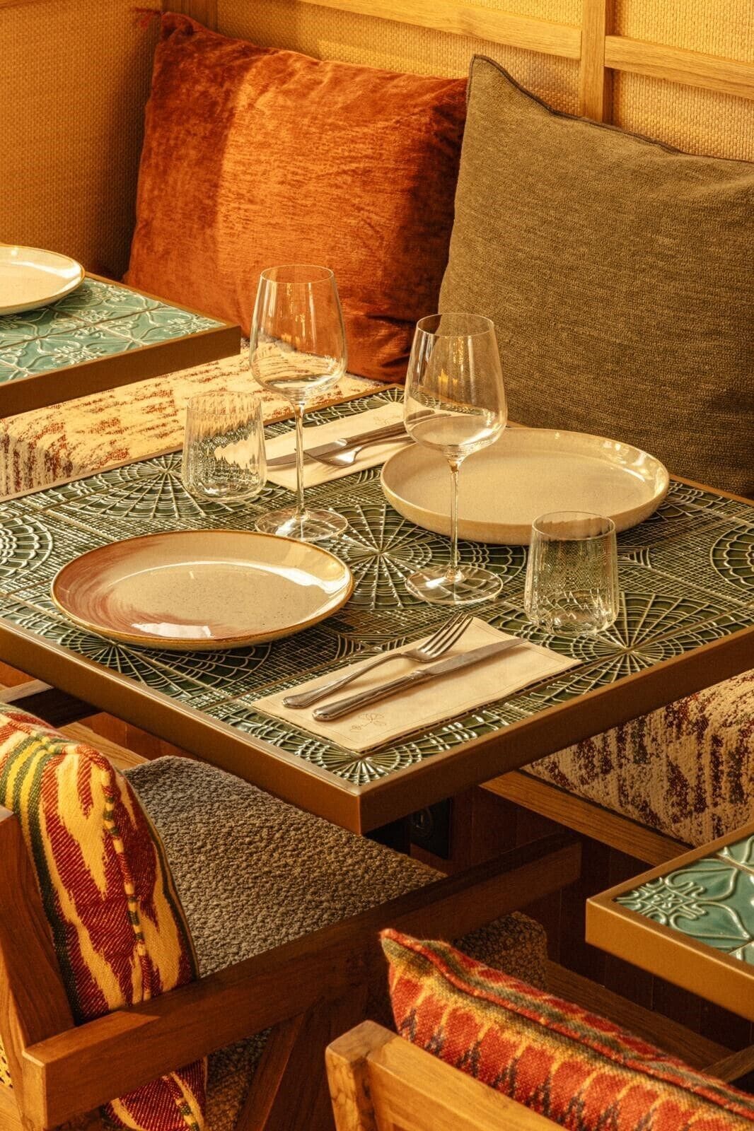 Uma área de refeições com uma mesa posta para dois, com toalha de mesa estampada, dois pratos, talheres e copos de vinho.