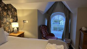 Quarto de hotel com cama, poltrona, luminária, janela com cortinas e vista externa.