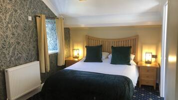 Quarto de hotel com cama, duas mesas de cabeceira com luminárias, janela com cortinas, radiador e papel de parede estampado.