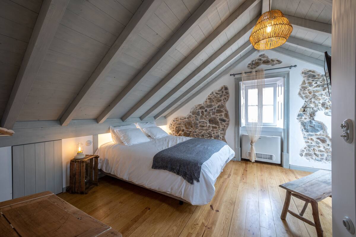 Quarto com teto de madeira, parede de pedra, cama com roupa de cama branca, mesinha pequena e luminária pendente.