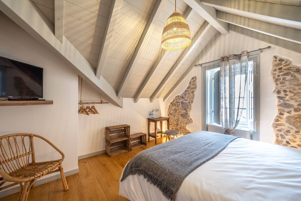 Quarto com cama, televisão, cadeira de vime, mesa pequena e parede de pedra.