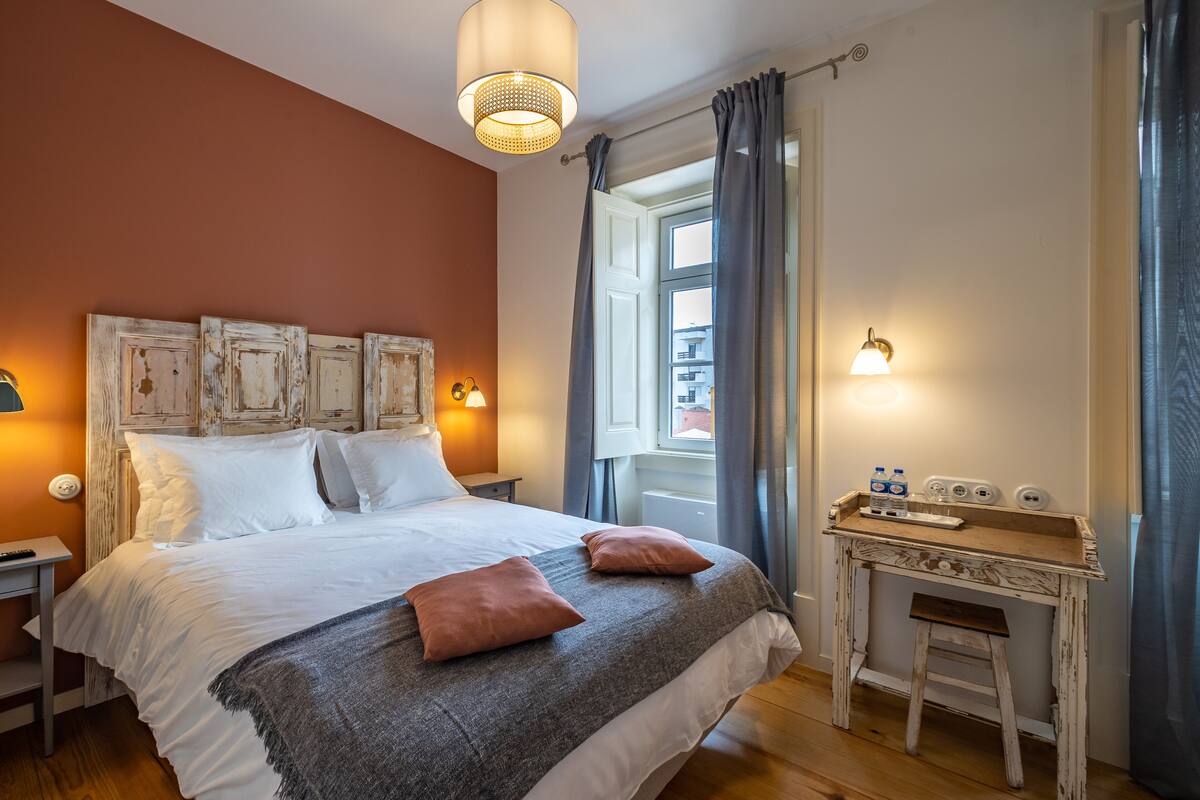 Uma cama bem arrumada com um cobertor cinzento e duas almofadas, uma escrivaninha de madeira com uma luminária e uma janela com cortinas.