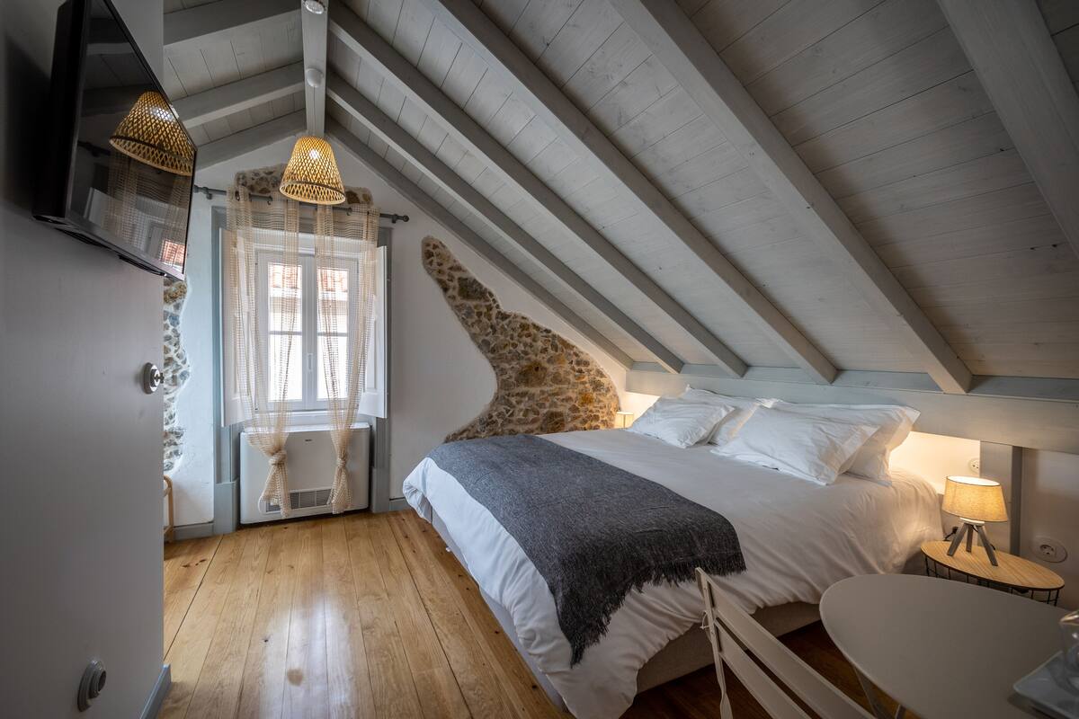 Quarto com teto inclinado, cama com lençóis brancos, mesa redonda pequena com abajur e parede de destaque em pedra.