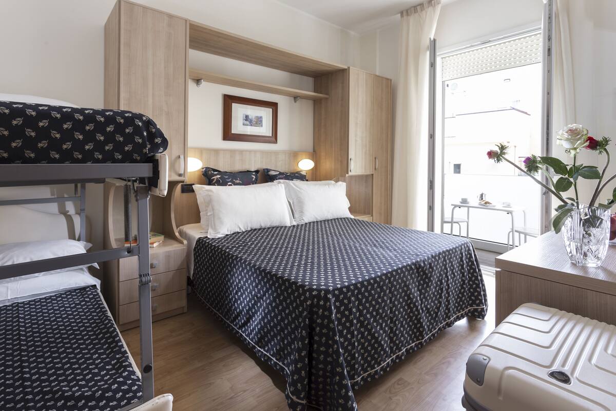 Quarto de hotel com beliche, cama de solteiro, escrivaninha com um vaso de flores e janela com vista.