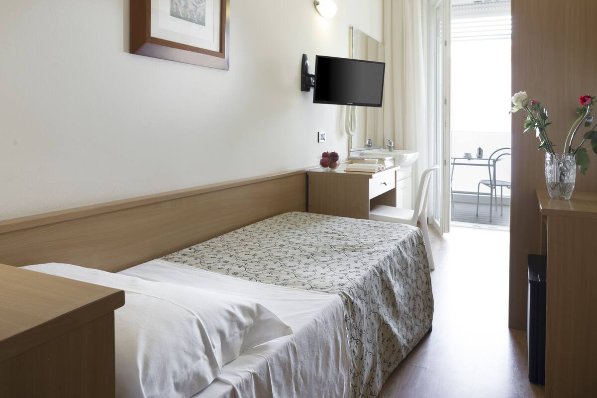 Quarto de hotel com cama, mesa de cabeceira, escrivaninha com cadeira, televisão na parede, banheiro com pia e espelho, e varanda com mesa e cadeiras.
