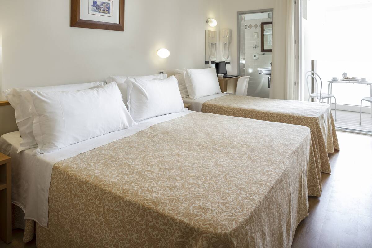Quarto de hotel com duas camas, uma escrivaninha, uma cadeira, uma mesinha e um banheiro visível por uma porta aberta.