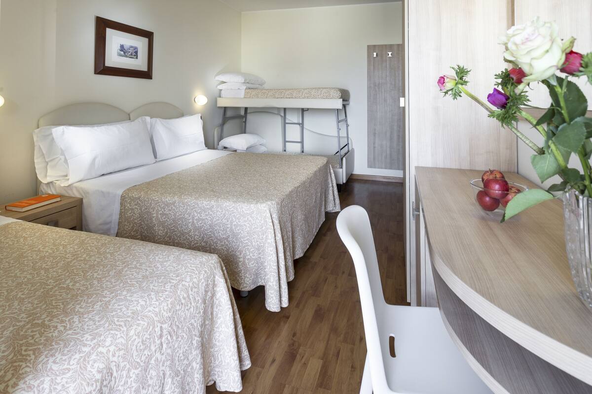 Quarto de hotel com duas camas, uma escrivaninha, uma cadeira e uma mesinha de centro com um vaso de flores.