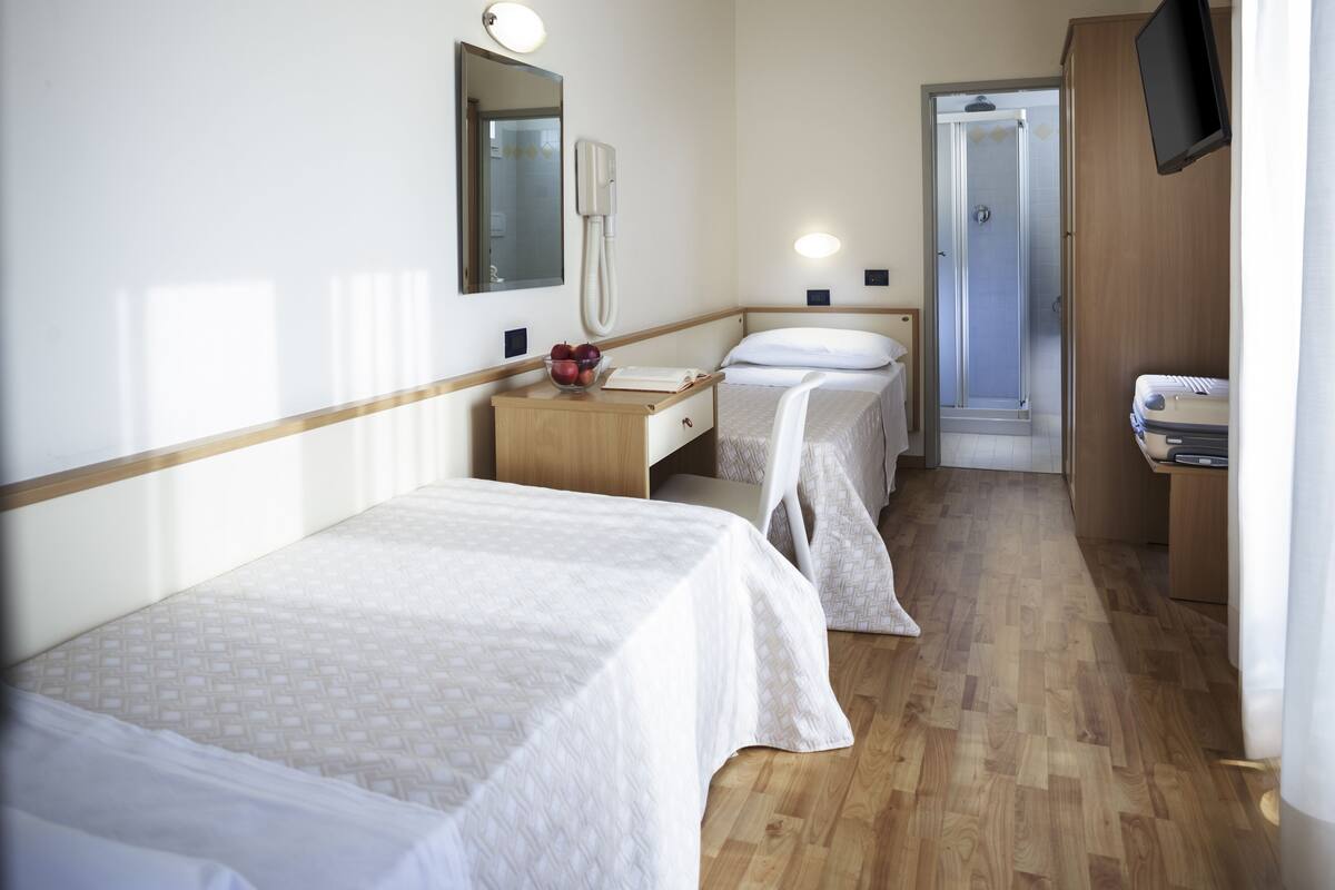 Quarto de hotel com duas camas de solteiro, mesa de cabeceira de madeira com uma tigela de frutas, luminária, espelho, TV fixada na parede e chuveiro.