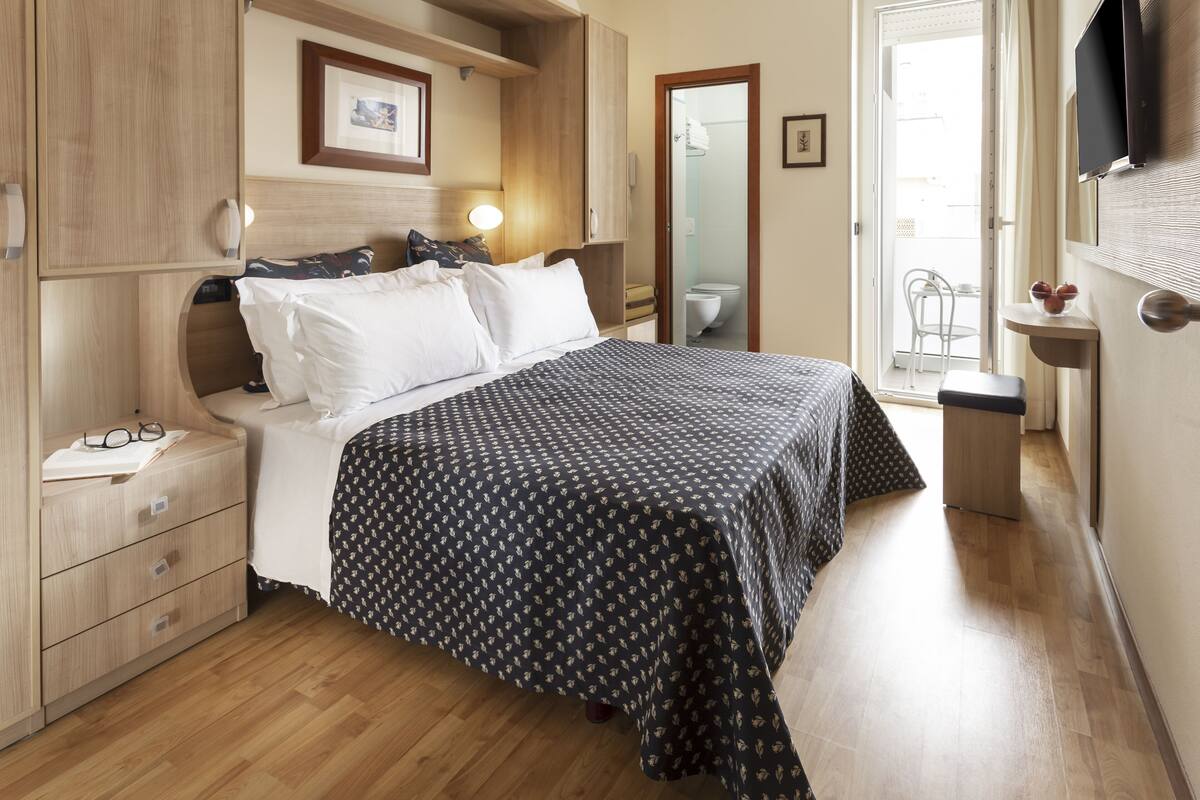 Quarto bem arrumado com cama, mesa de cabeceira e uma pequena escrivaninha. Há uma porta que dá para o banheiro e uma televisão na parede.
