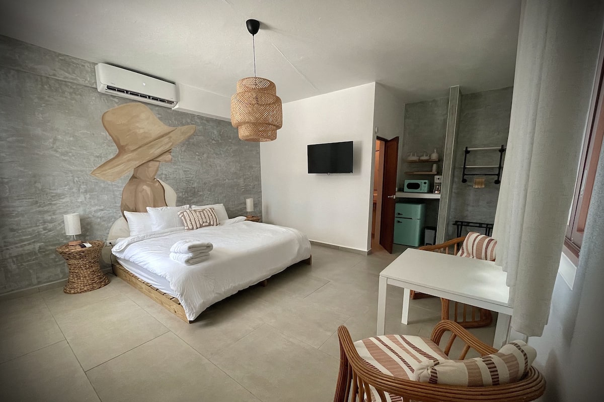 Quarto moderno com uma cama grande, área de refeições com móveis de vime, televisão e uma cozinha compacta.
