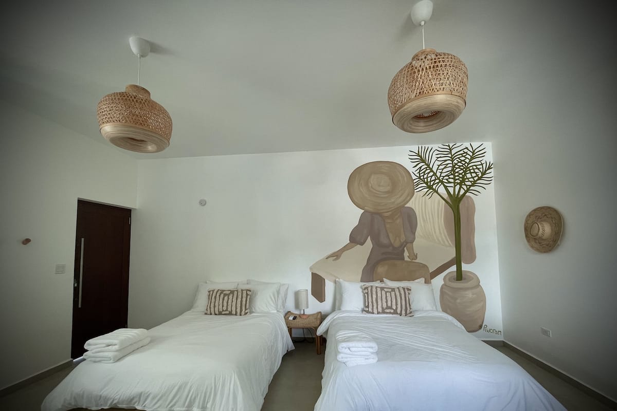 Quarto com duas camas de solteiro, porta de madeira e uma pintura na parede retratando uma mulher com chapéu.