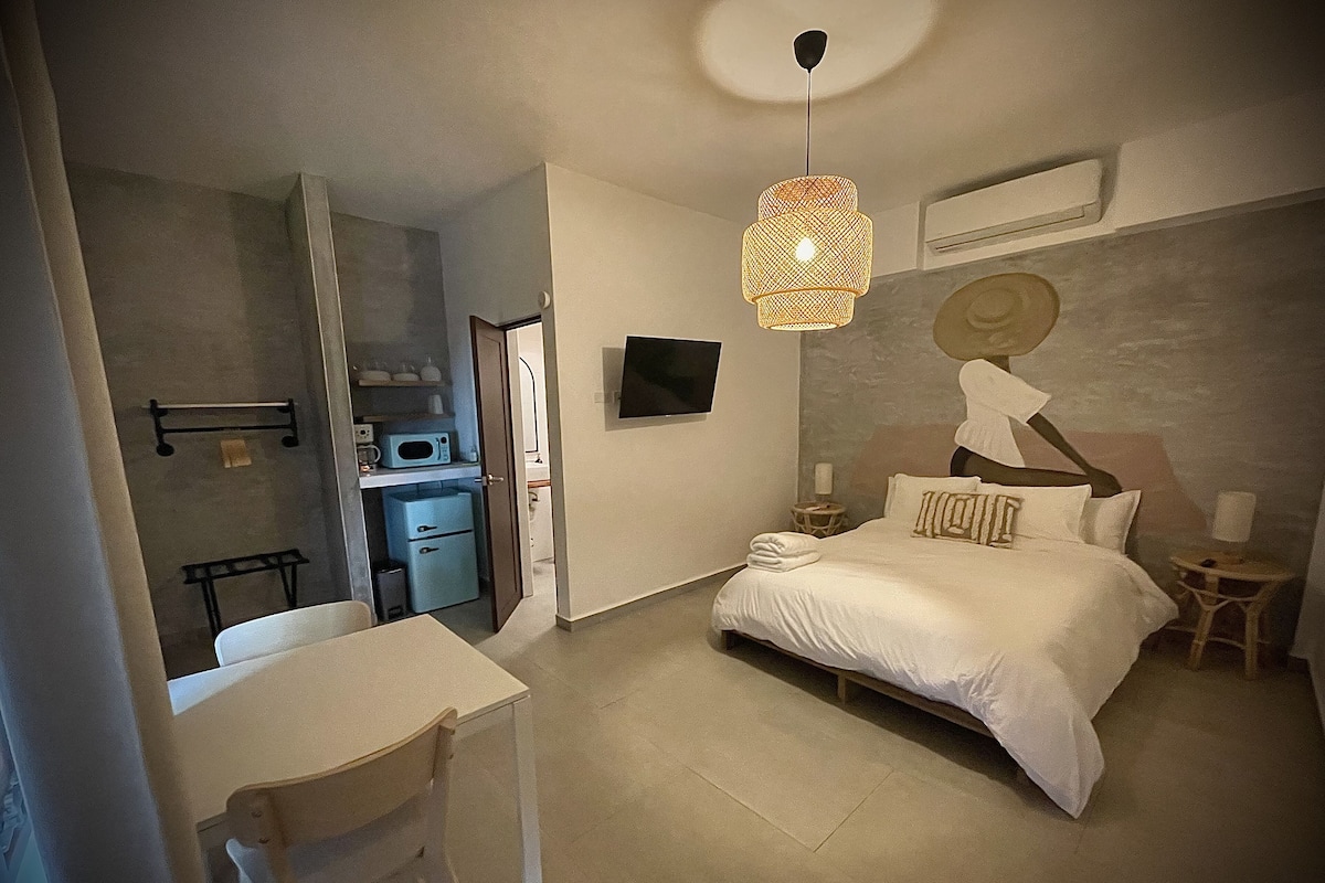Quarto com cama, mesas de cabeceira, escrivaninha, cadeira, televisão e micro-ondas na cozinha compacta adjacente.